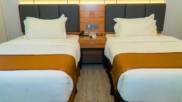 Quarto de hotel com duas camas, uma mesa de cabeceira com telefone e controle remoto, e cabeceira de madeira com interruptor de luz.