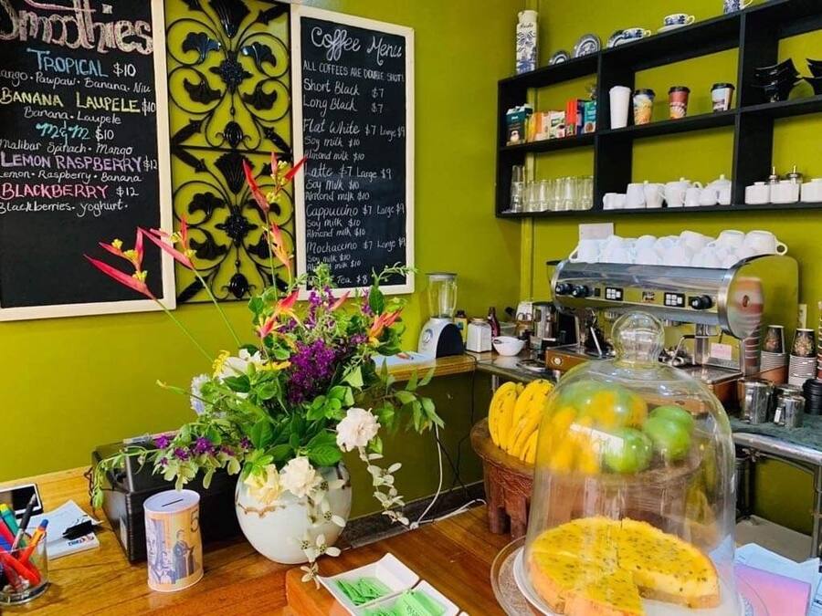 Balcão de café com um quadro de menu, um registrador de caixa e uma exposição de bananas e uma tigela de frutas tampada.