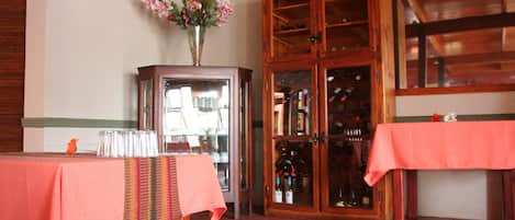 Um quarto com duas mesas cobertas por toalhas vermelhas, um armário de madeira com portas de vidro e um vaso com flores numa prateleira.