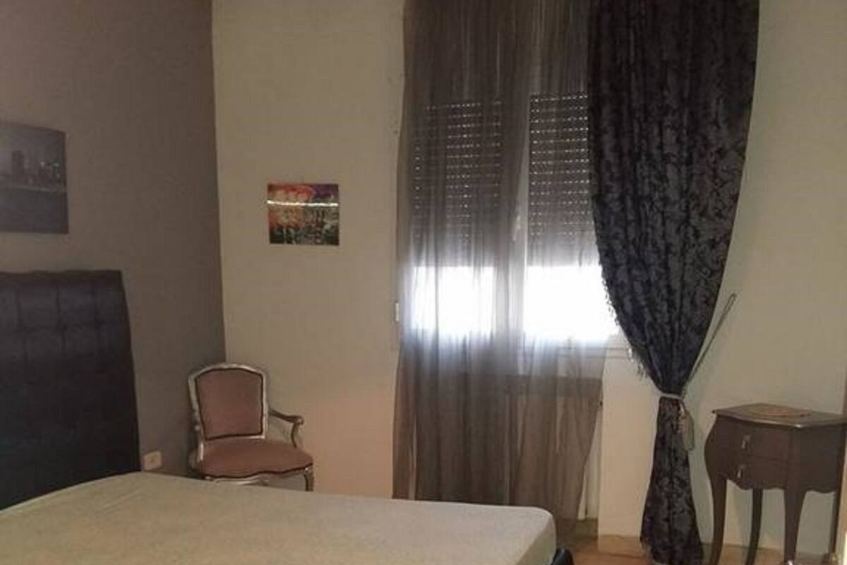 Quarto com uma cama, uma cadeira, uma mesa pequena e uma janela com cortinas.