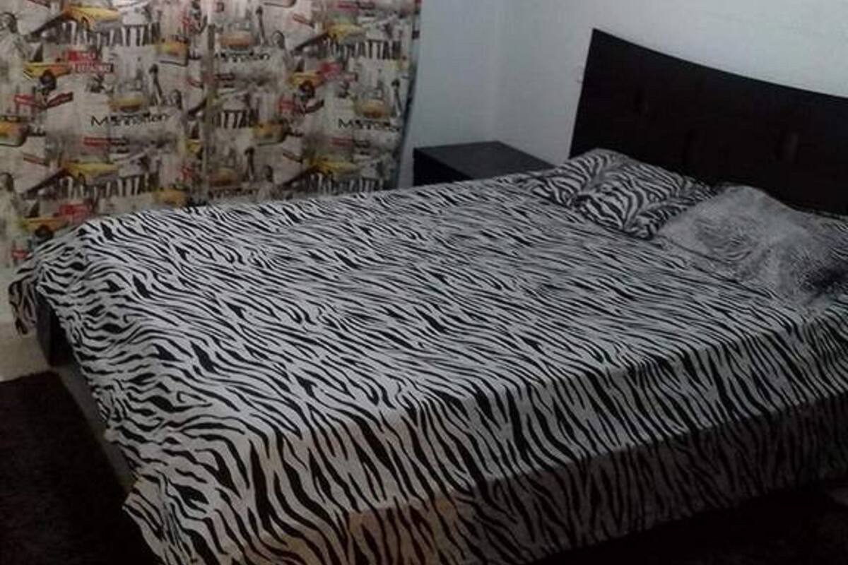 Quarto com colcha estampada em padrão de zebra, mesa de cabeceira preta, luminária fixada na parede e pôster de um carro na parede.
