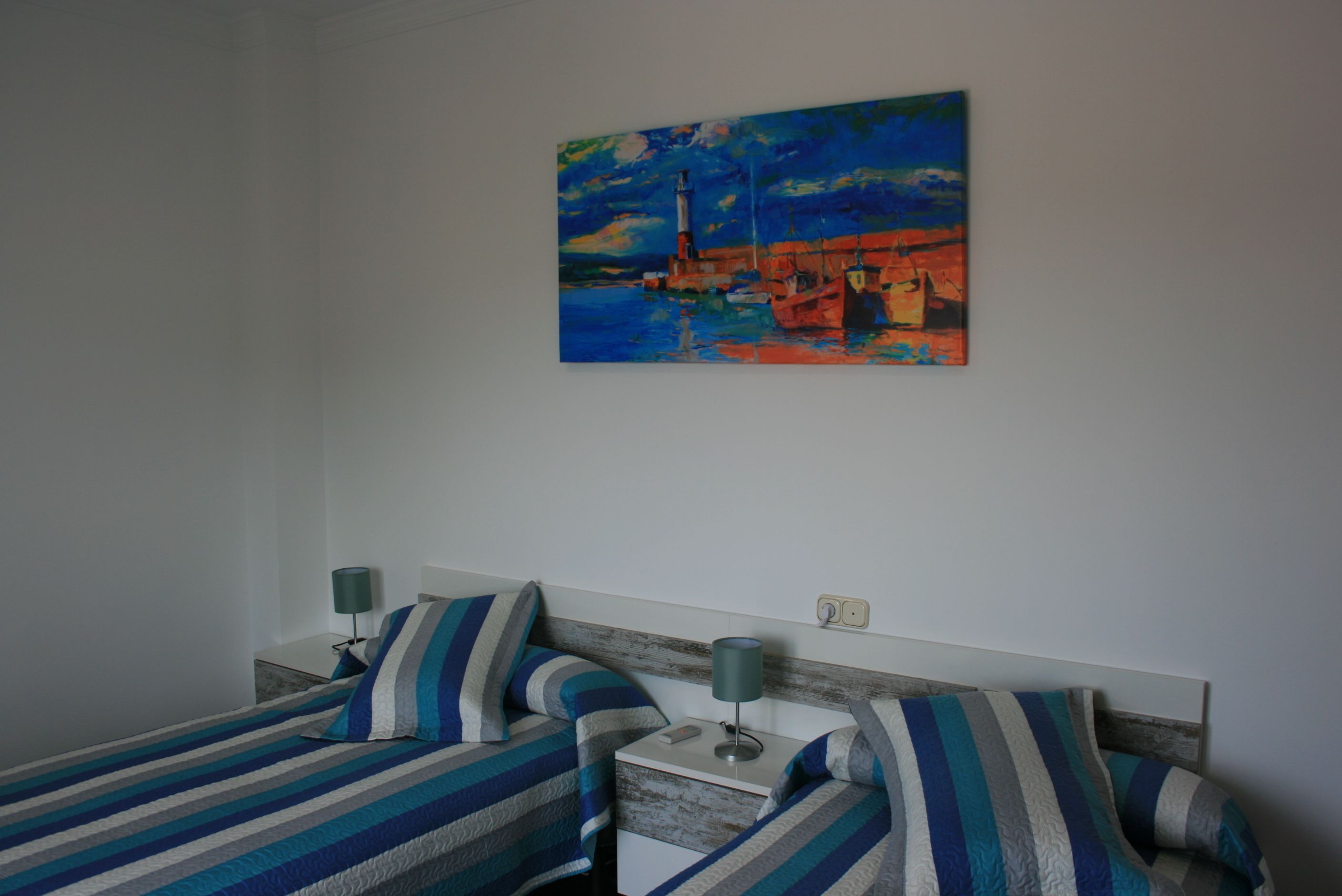 Quarto de hotel com duas camas, uma mesa de cabeceira, um abajur e um quadro representando uma paisagem costeira.