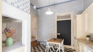 Uma cozinha com chão xadrez, mesa de madeira, cadeiras brancas, luminária pendente e porta com padrão azul e branco.