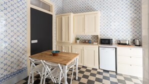 Uma cozinha compacta com uma mesa de madeira e cadeiras brancas, micro-ondas, chaleira e um frigorífico pequeno.