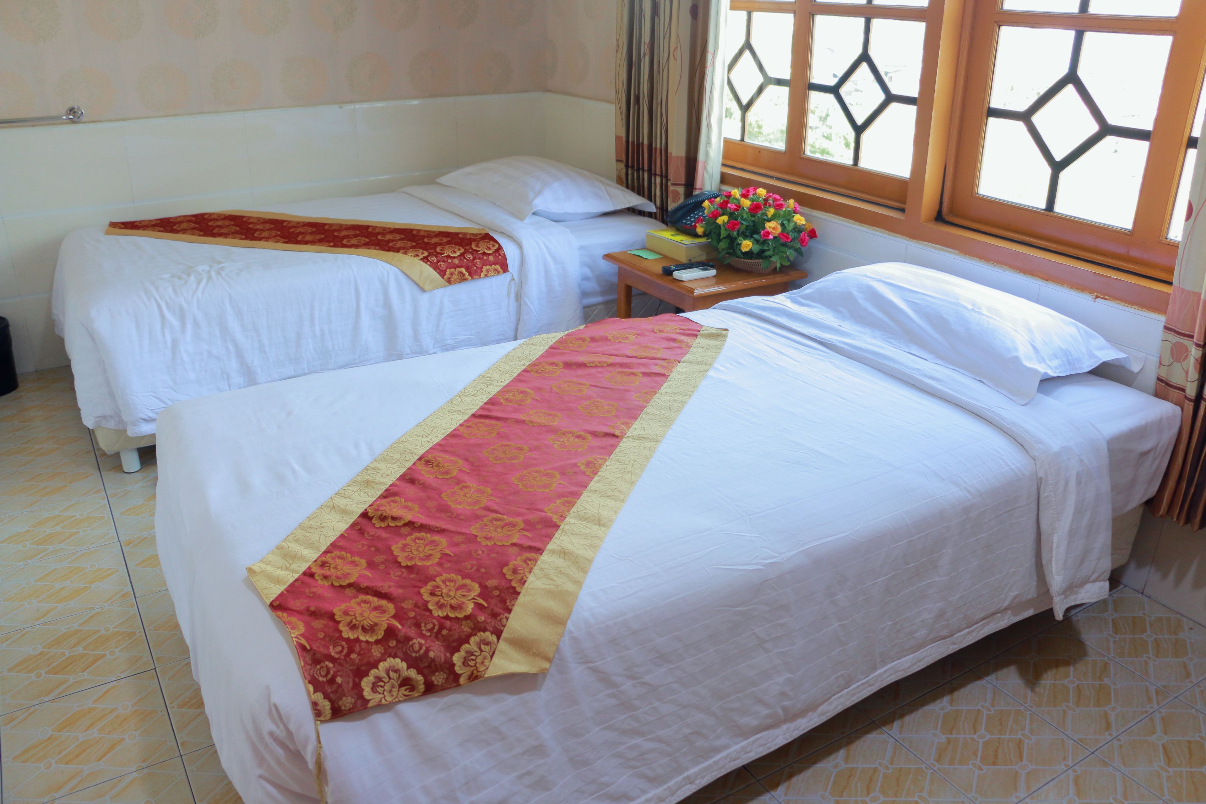 Quarto de hotel com duas camas, uma mesinha de cabeceira com um vaso de flores e uma janela com padrão geométrico.