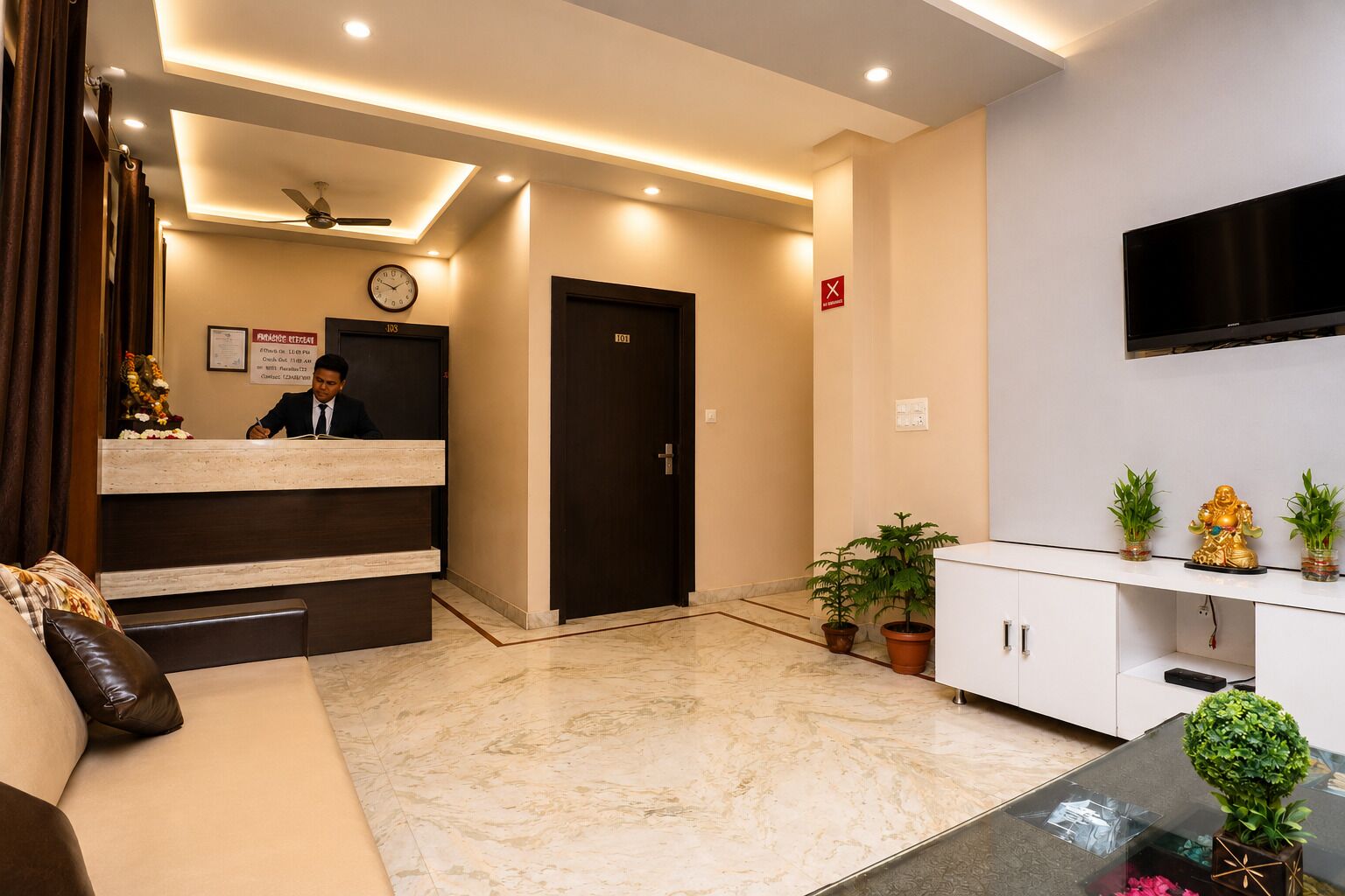 Uma área de recepção de hotel com balcão, relógio, porta fechada, televisão na parede e um espaço de estar com sofá e mesa de centro.