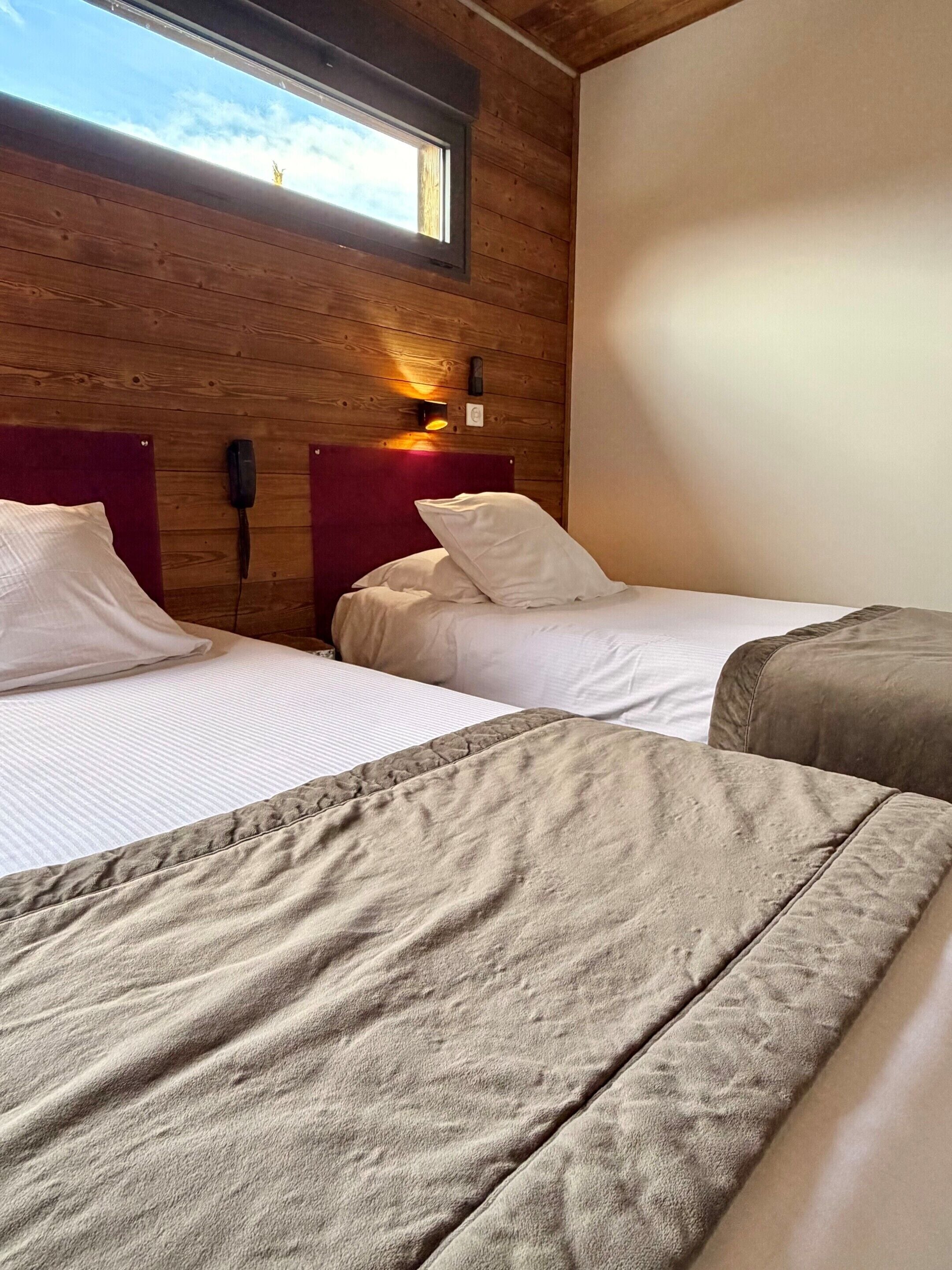 Quarto com duas camas, paredes de madeira, uma janela e luminária fixada na parede.