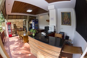 Uma área de recepção com balcão de madeira, geladeira Coca-Cola, caixa eletrônico e um computador.