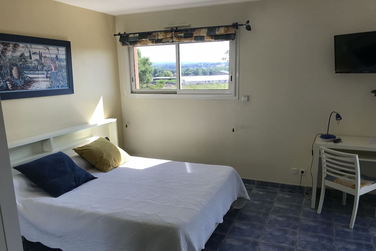 Quarto com cama, escrivaninha, cadeira, janela com vista e televisão na parede.