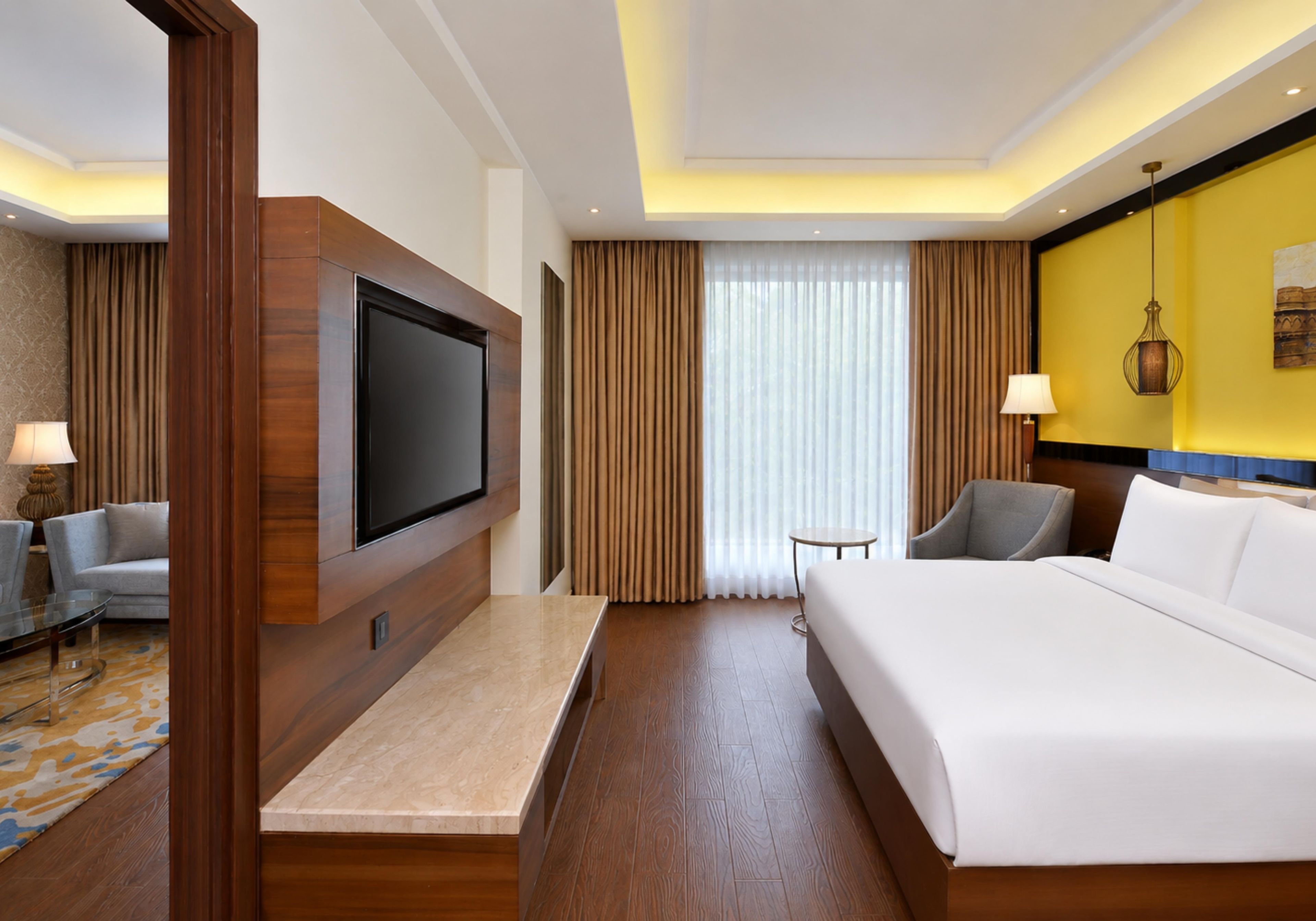 Quarto de hotel com uma cama grande, televisão de tela plana na parede, área de estar com uma poltrona e uma mesa pequena, e uma janela ampla com cortinas.
