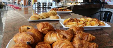 Uma mesa de buffet com diversos doces e produtos de panificação, incluindo croissants e tartes de fruta.
