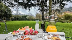 Disposição para café da manhã ao ar livre com pães, frutas e bebidas sobre uma mesa em área gramada.