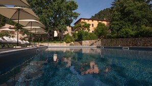 Piscina com espreguiçadeiras e guarda-sóis, cercada por árvores e uma parede de pedra.