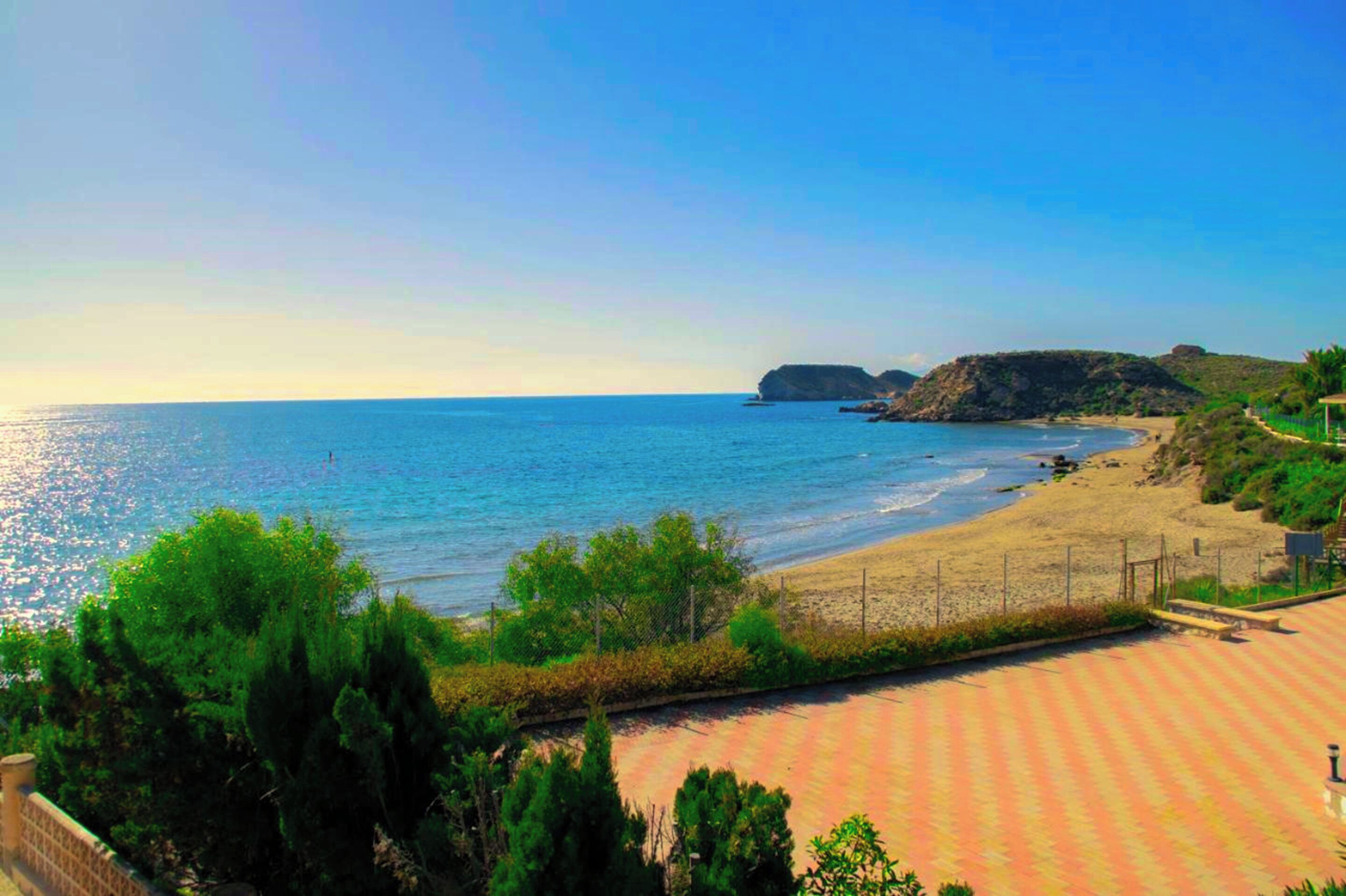 Uma praia com calçada pavimentada, afloramento rochoso e vista clara do mar.