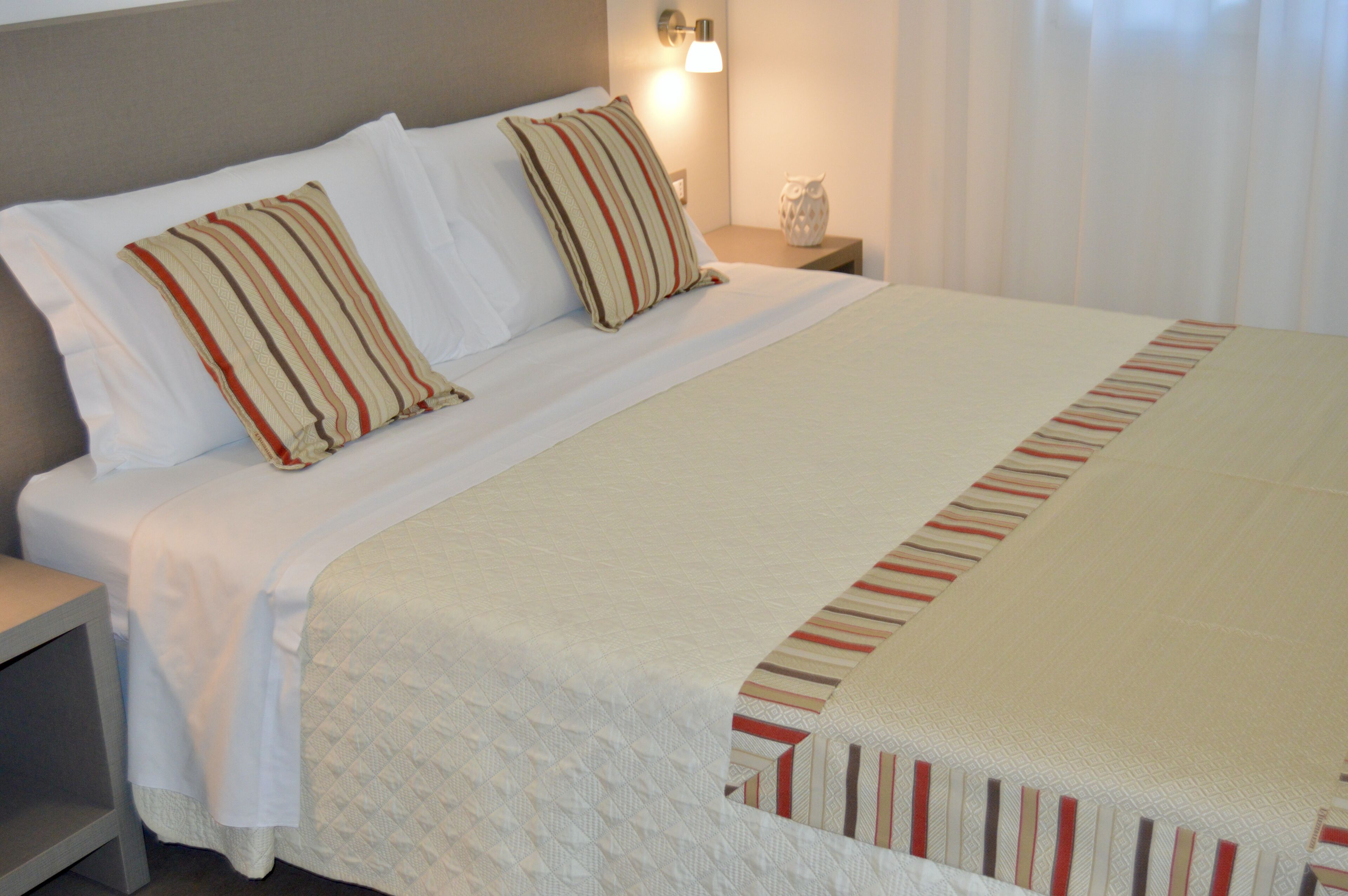 Uma cama bem arrumada com lençóis brancos e almofadas listradas, uma mesinha de cabeceira e um abajur fixado na parede.
