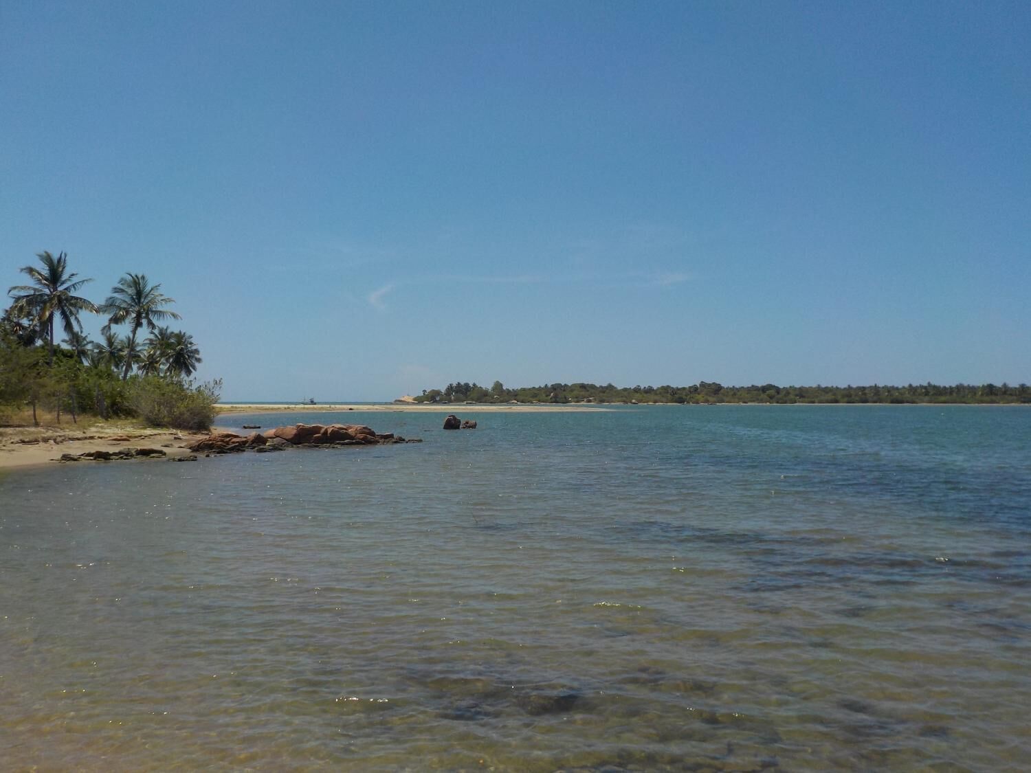 Uma praia com águas claras e rasas, palmeiras e uma linha de costa ao longe.