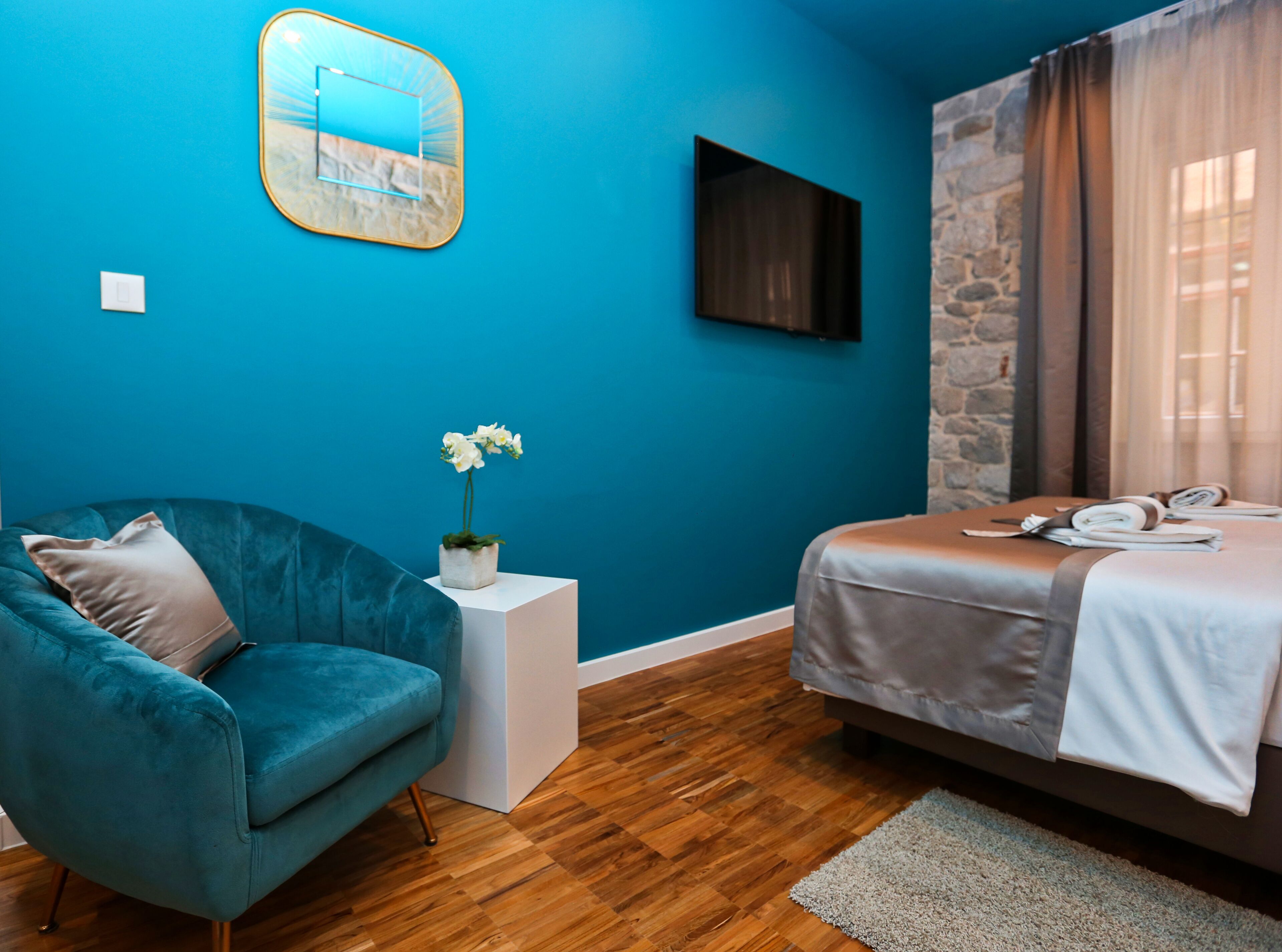 Quarto com poltrona azul-esverdeada, mesinha de apoio branca com uma orquídea em vaso, cama com colcha branca, parede de pedra e televisão montada.