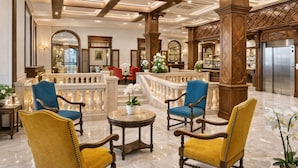 Um hall amplo com piso de mármore, cadeiras de madeira clássicas, uma mesa redonda central e uma área de recepção com balcão e cadeiras.