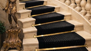 Uma escadaria imponente com degraus revestidos de carpete preto, um candelabro dourado e uma placa indicando a recepção.