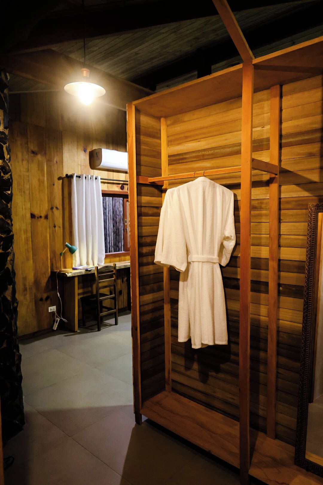 Quarto de madeira com roupão pendurado em um suporte, uma mesa com abajur e uma janela com cortinas.