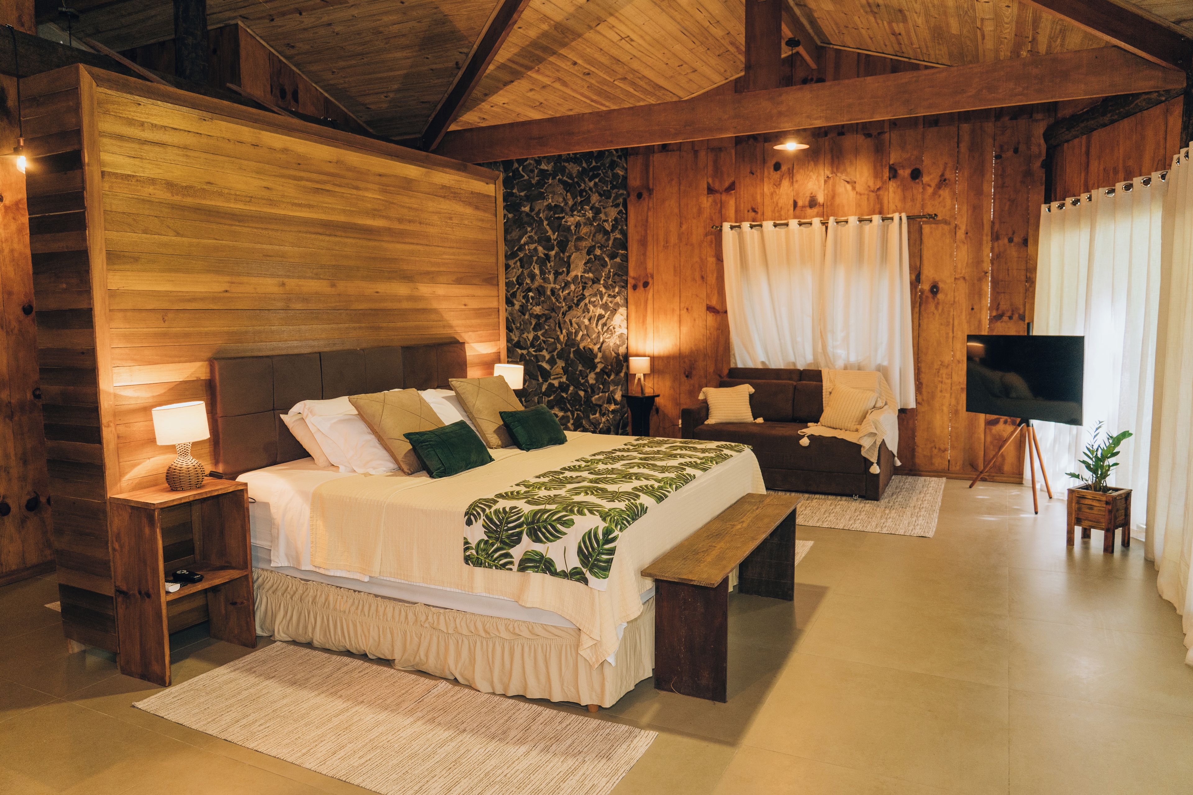 Quarto com decoração em madeira, com cama, mesas de cabeceira, televisão e parede de pedra.