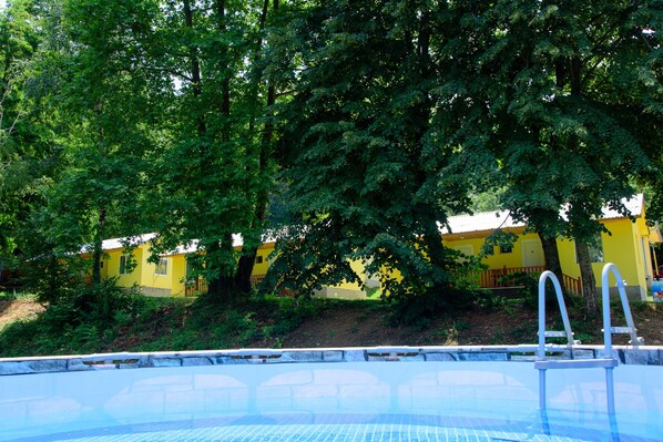 Piscina com superfície de água azul cristalina, rodeada por árvores verdes exuberantes e um edifício amarelo ao fundo.