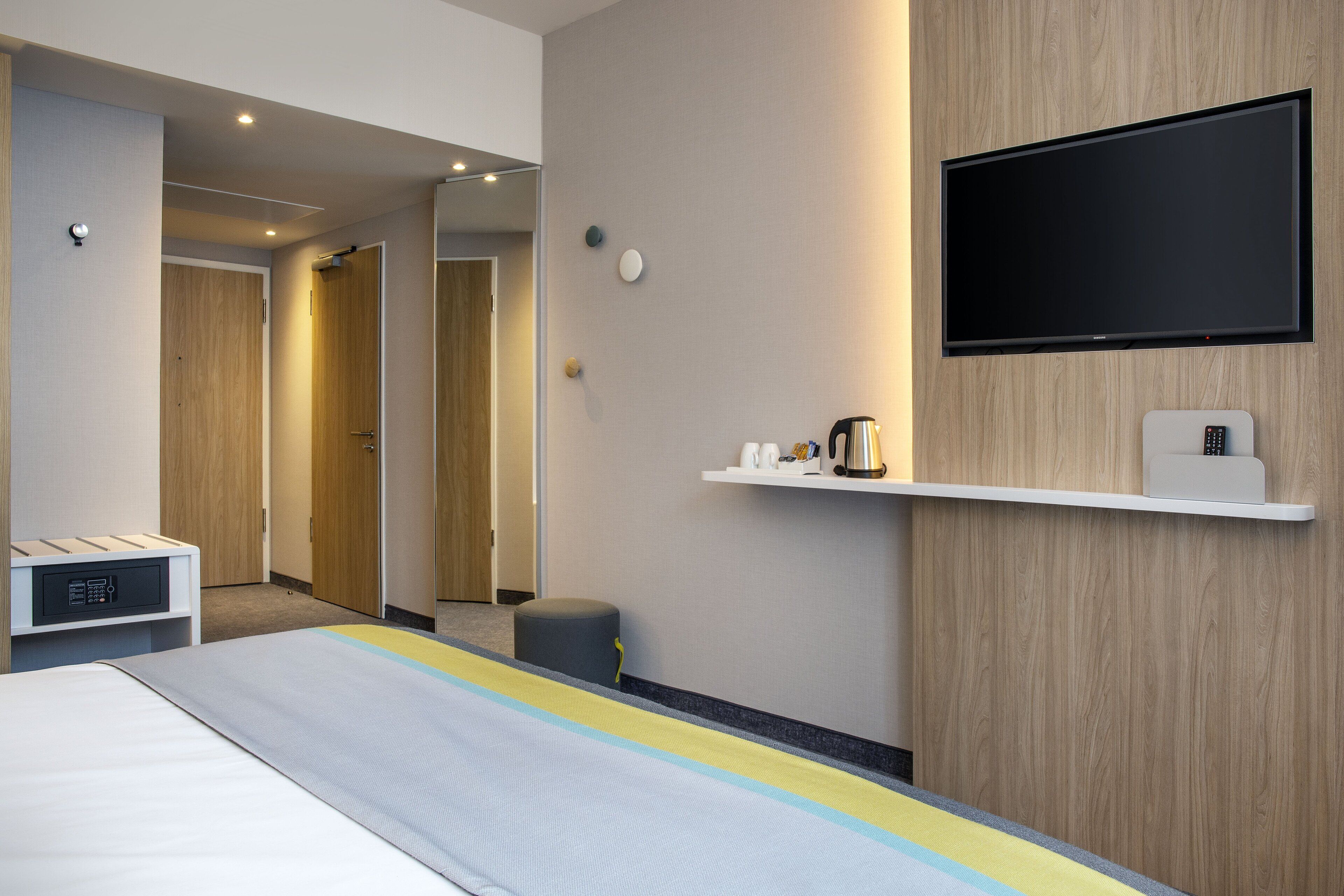 Quarto de hotel moderno com cama, televisão de tela plana na parede, prateleira pequena com cafeteira e painel de controle.