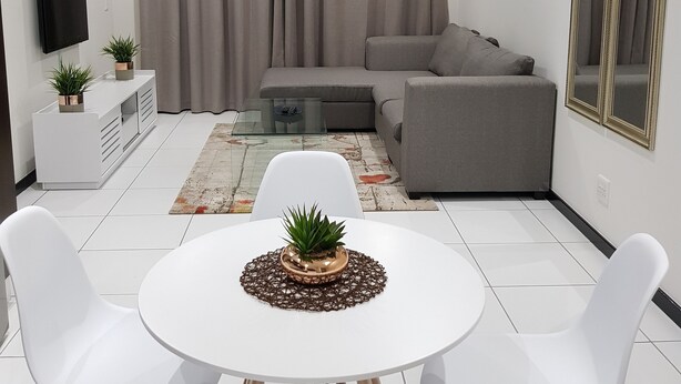 Uma sala de estar moderna com um sofá cinzento, uma mesa de jantar branca com duas cadeiras, uma mesa de centro de vidro e um armário branco com uma televisão de tela plana.