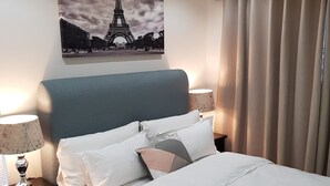 Cama bem arrumada com lençóis brancos e várias almofadas, luminária de cabeceira, controle remoto e quadro com a Torre Eiffel na parede.