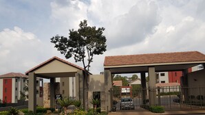 Uma entrada com portão e área de espera coberta, céu claro e edifícios ao fundo.