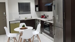 Uma cozinha moderna com armários escuros, eletrodomésticos de aço inoxidável e um espaço de refeições com cadeiras brancas e uma mesa pequena.