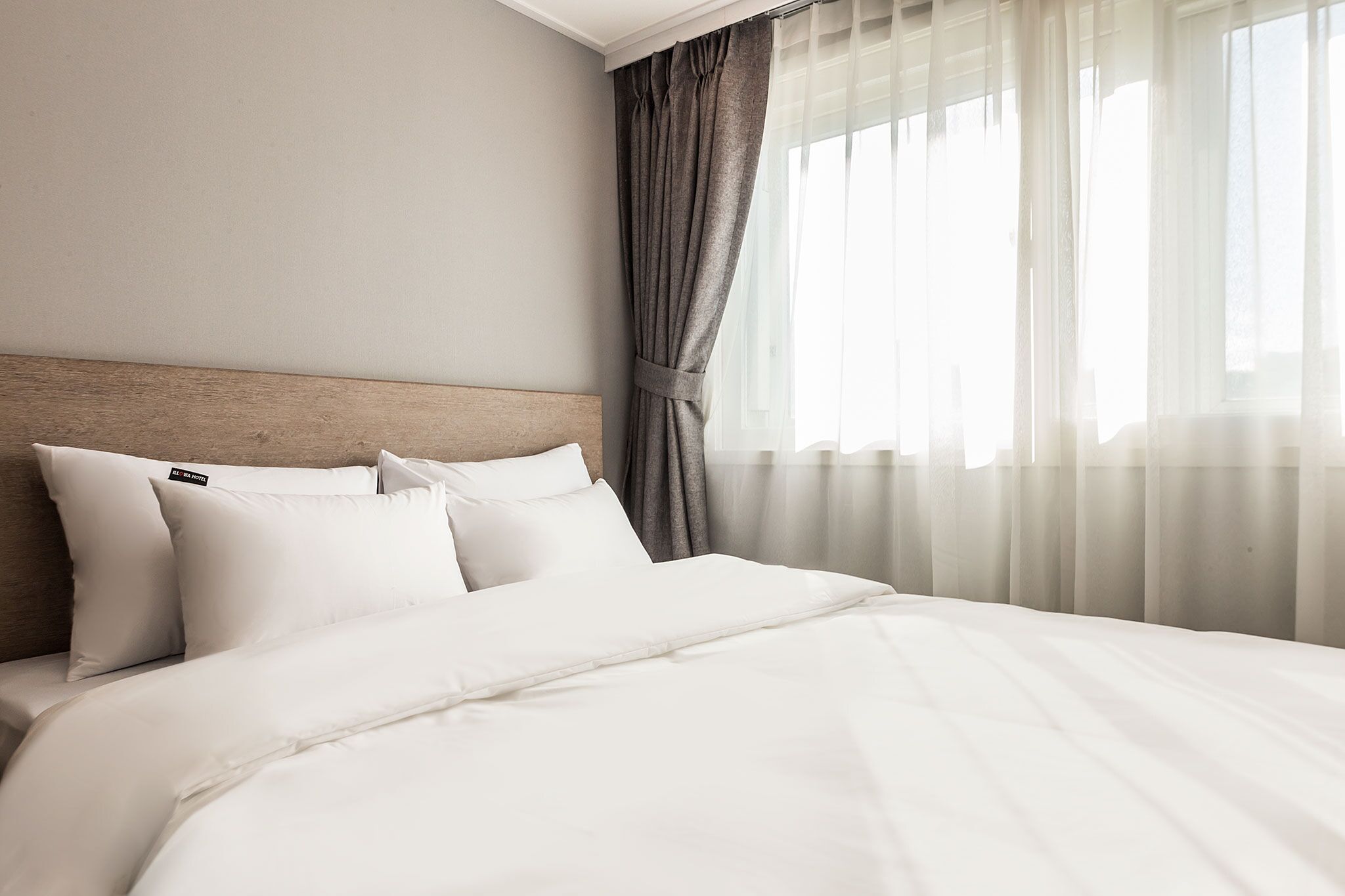 Cama bem arrumada com lençóis e almofadas brancos, cabeceira de madeira e cortinas leves na janela.