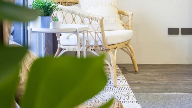 Uma cadeira de vime com almofada, uma mesa pequena com uma planta e um enfeite de macramé num quarto com janela e tapete estampado.