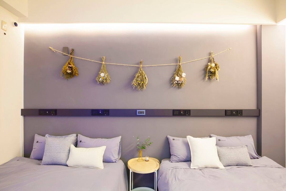 Quarto de hotel moderno com duas camas, cada uma com cabeceira, mesa de cabeceira pequena e luminária fixada na parede.