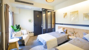 Quarto de hotel com duas camas, área de estar com uma mesa, uma janela grande com cortinas e uma porta que dá para outro quarto.