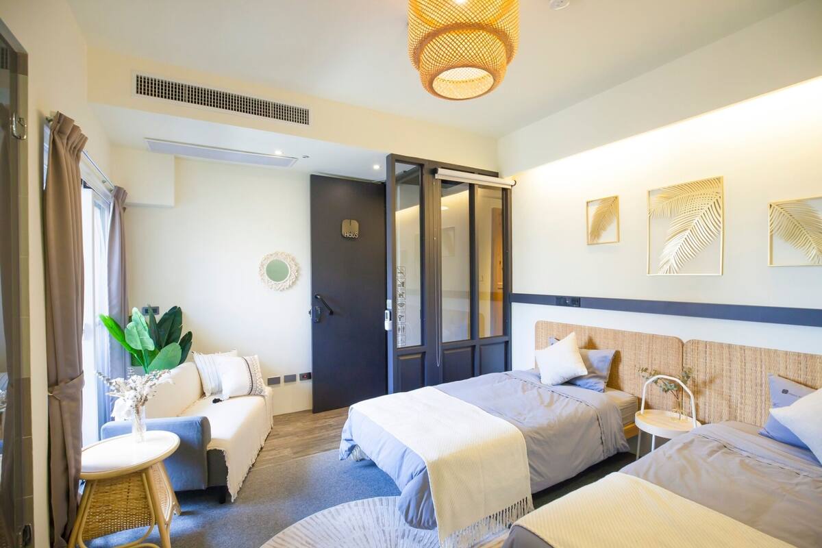 Quarto de hotel com duas camas, área de estar com uma mesa, uma janela grande com cortinas e uma porta que dá para outro quarto.
