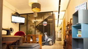Um hall de hotel moderno com um tapete azul escrito 'oh, Hello', uma máquina de costura antiga, uma cadeira vermelha, um patinete e uma escada.