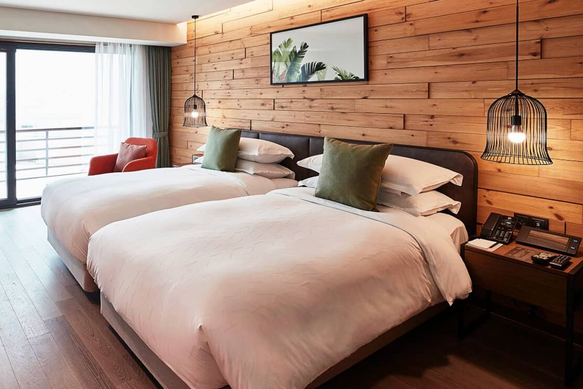 Quarto de hotel com duas camas, uma escrivaninha, uma cadeira, parede de madeira e janela com cortinas.