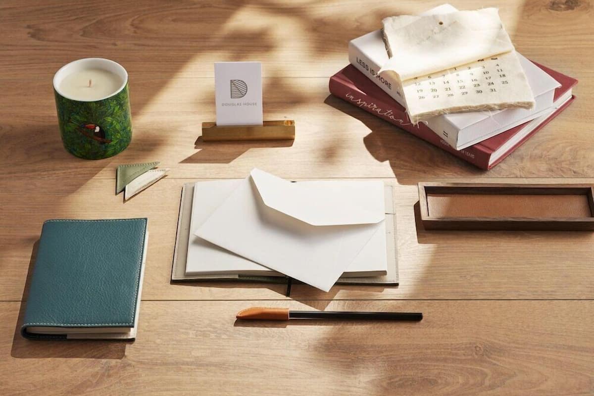 Uma mesa de madeira com uma vela verde, um cartão de visita, uma pilha de livros, um caderno, uma caneta e uma pequena caixa.