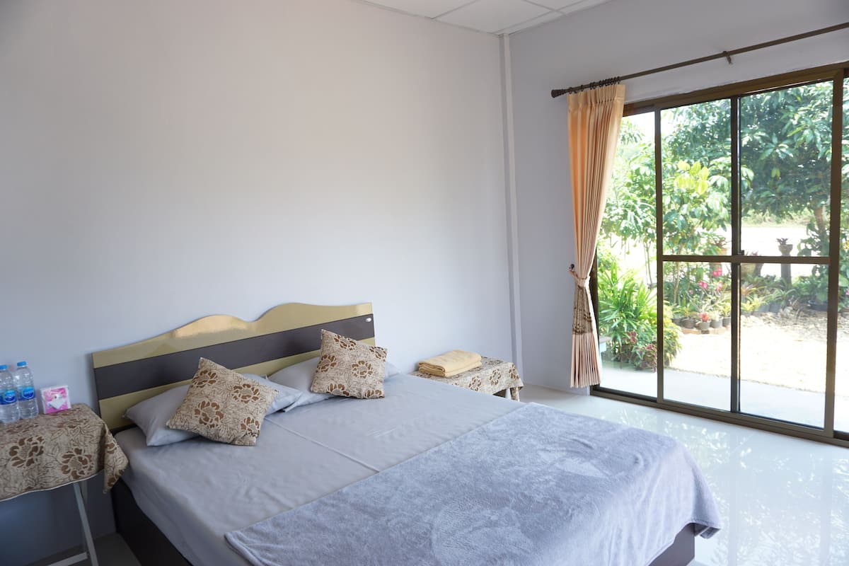 Cama bem arrumada com colcha cinza e duas almofadas estampadas, mesa de cabeceira com água engarrafada e um objeto rosa, e janela com cortina que dá para um jardim.
