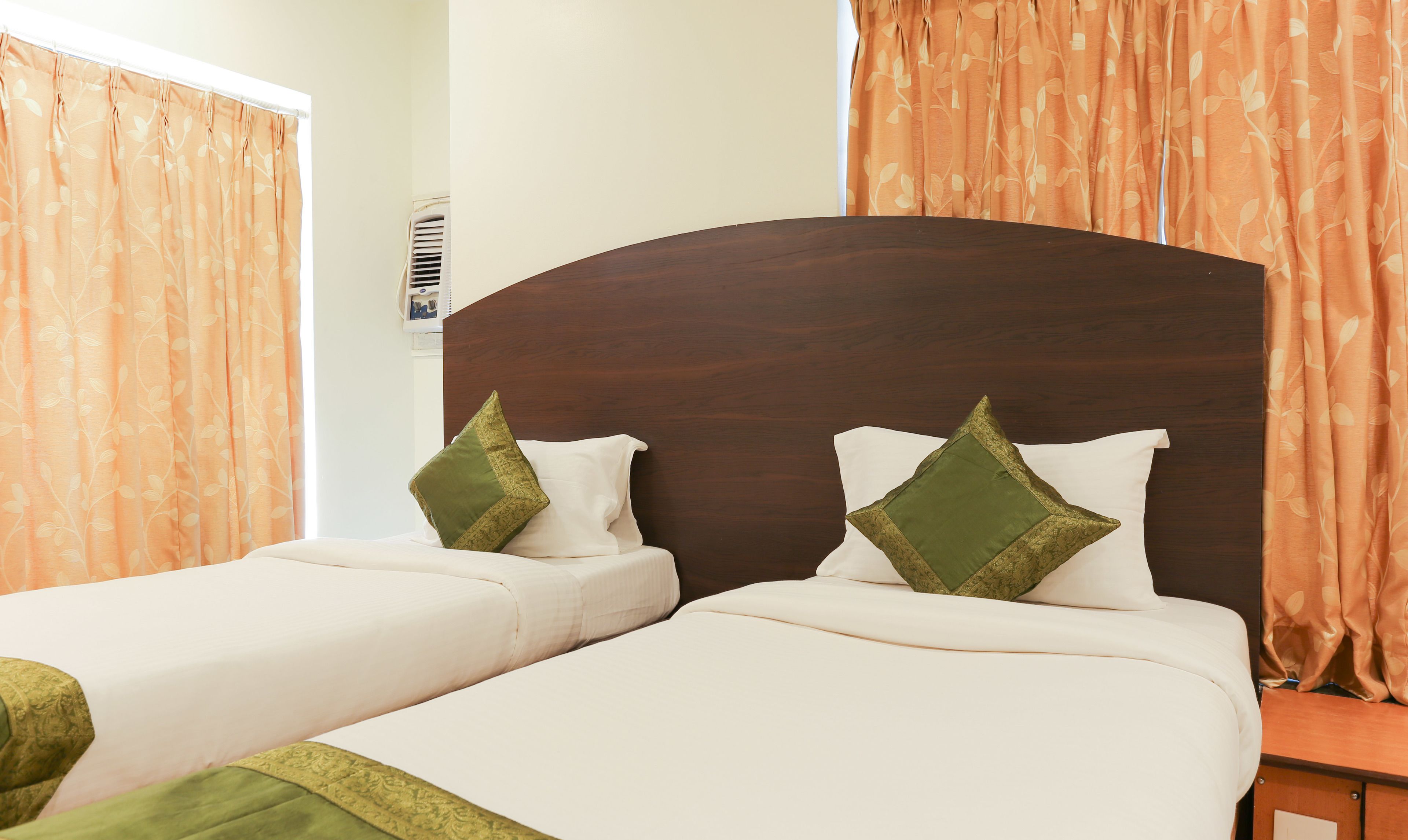 Quarto de hotel com duas camas de solteiro, cada uma com cabeceira de madeira, roupa de cama branca e almofadas decorativas verdes.