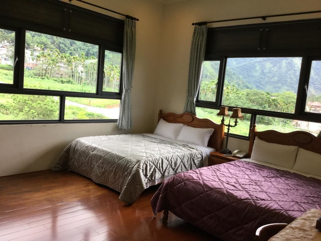 Quarto com duas camas, janelas amplas, piso de madeira e vista para o verde.