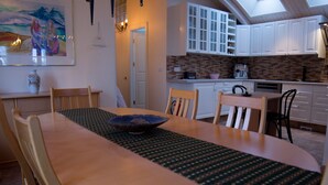 Uma área de refeições com mesa de madeira, cadeiras e uma tigela decorativa. Cozinha ao fundo com armários brancos e painel de backsplash de tijolos.