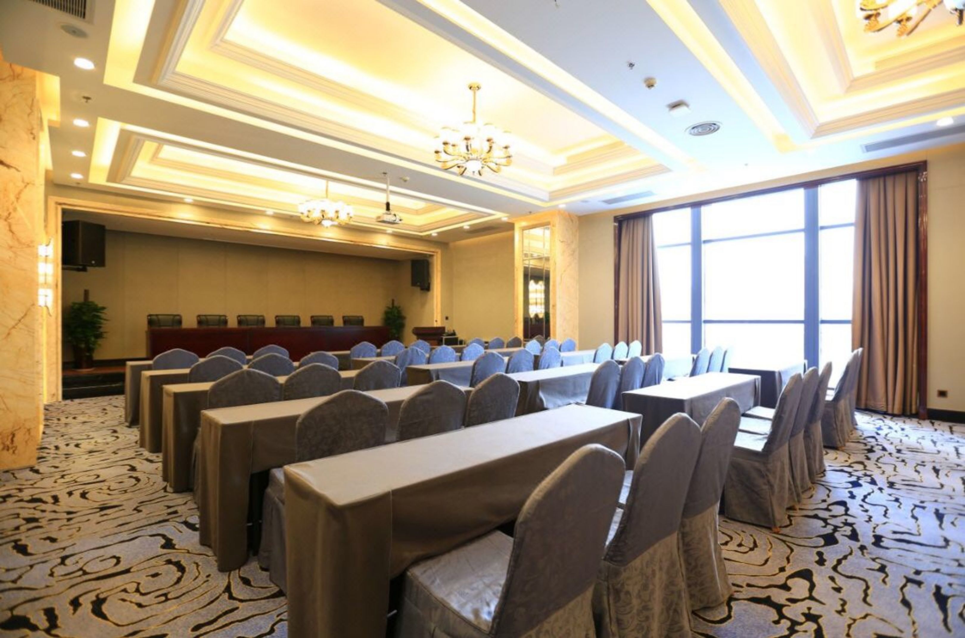 Uma sala de conferências com fileiras de mesas e cadeiras, um candelabro e amplas janelas com cortinas.