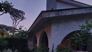 Um edifício com janelas em arco e telhado plano, rodeado por vegetação exuberante e plantas floridas.