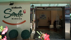 A entrada de um hotel chamado 'Smile Mansion', com uma placa de boas-vindas, porta de vidro e vista do interior, incluindo uma área de estar e uma parede com obras de arte.