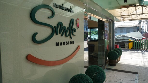 Entrada de um hotel com uma placa indicando "Smile Mansion" e plantas em vasos na frente.