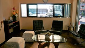 Quarto com mesa de centro de vidro, duas poltronas, um sofá, uma mesinha lateral e janela com vista para a rua.