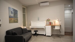 Um espaço de estar compacto com um sofá cinzento, uma secretária branca com uma cadeira e um banquinho pequeno.