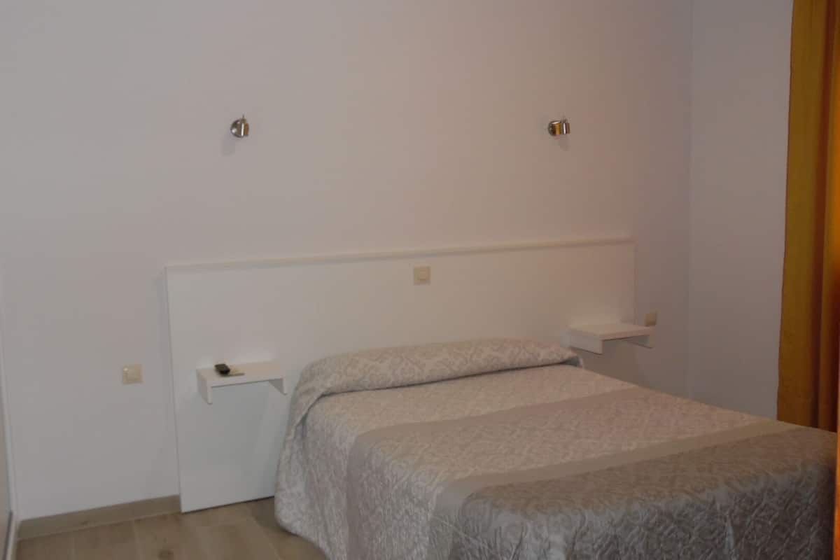 Quarto pequeno e simples, com cama, mesa de cabeceira e prateleira na parede.